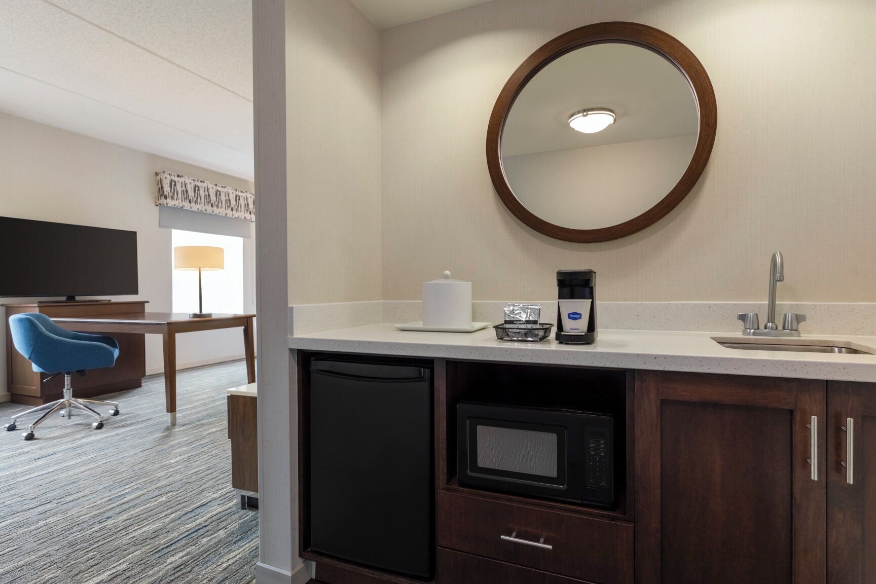 Quarto de hotel com escrivaninha, cadeira, micro-ondas, máquina de café e um espelho grande.