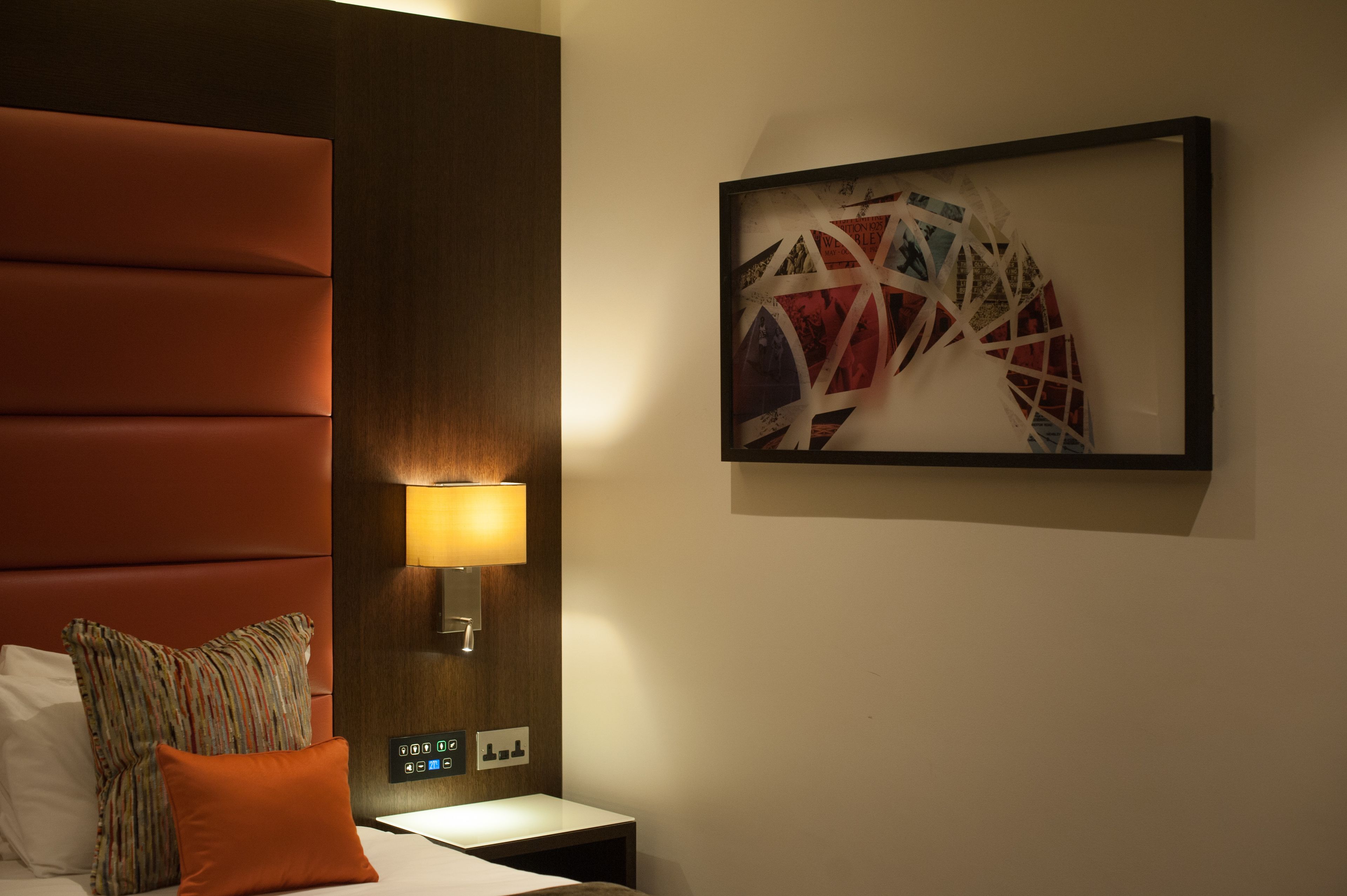 Quarto de hotel com cama, mesa de cabeceira, luminária fixada na parede e pintura abstrata emoldurada.