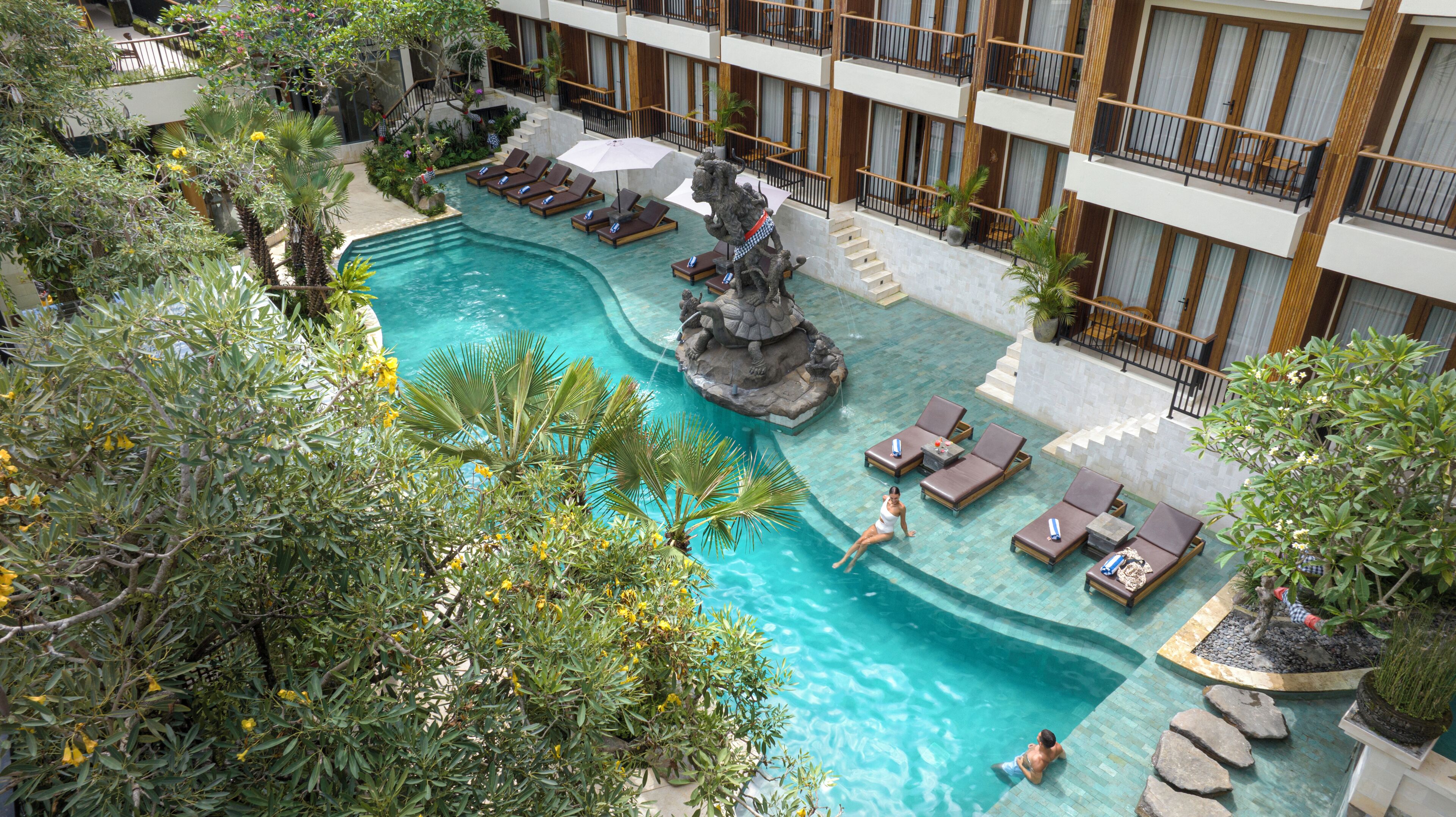 Uma área de piscina de hotel com uma fonte central, espreguiçadeiras e vista para as varandas do edifício.