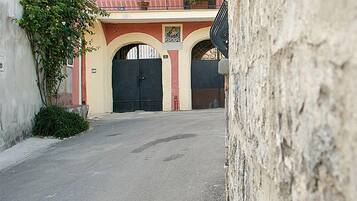 Uma rua estreita com um edifício de fachada vermelha, duas grades pretas e uma pequena varanda com plantas.