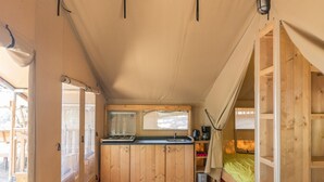 Cozinha compacta com armários de madeira, pia e fogão. Há uma cama com cobertor verde e uma garrafa na mesa.