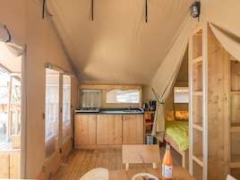 Cozinha compacta com armários de madeira, pia e fogão. Há uma cama com cobertor verde e uma garrafa na mesa.