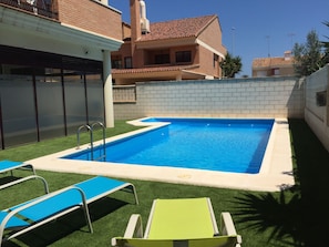 Piscina retangular com interior azul, cercada por grama sintética e uma parede de tijolos brancos. Há duas cadeiras de descanso, uma azul e outra verde, posicionadas sobre a grama.