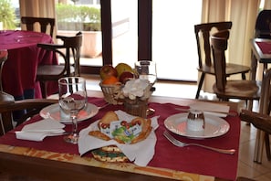 Uma mesa de jantar posta com uma cesta de pão, um copo de água, um prato com sobremesa e uma fruteira.