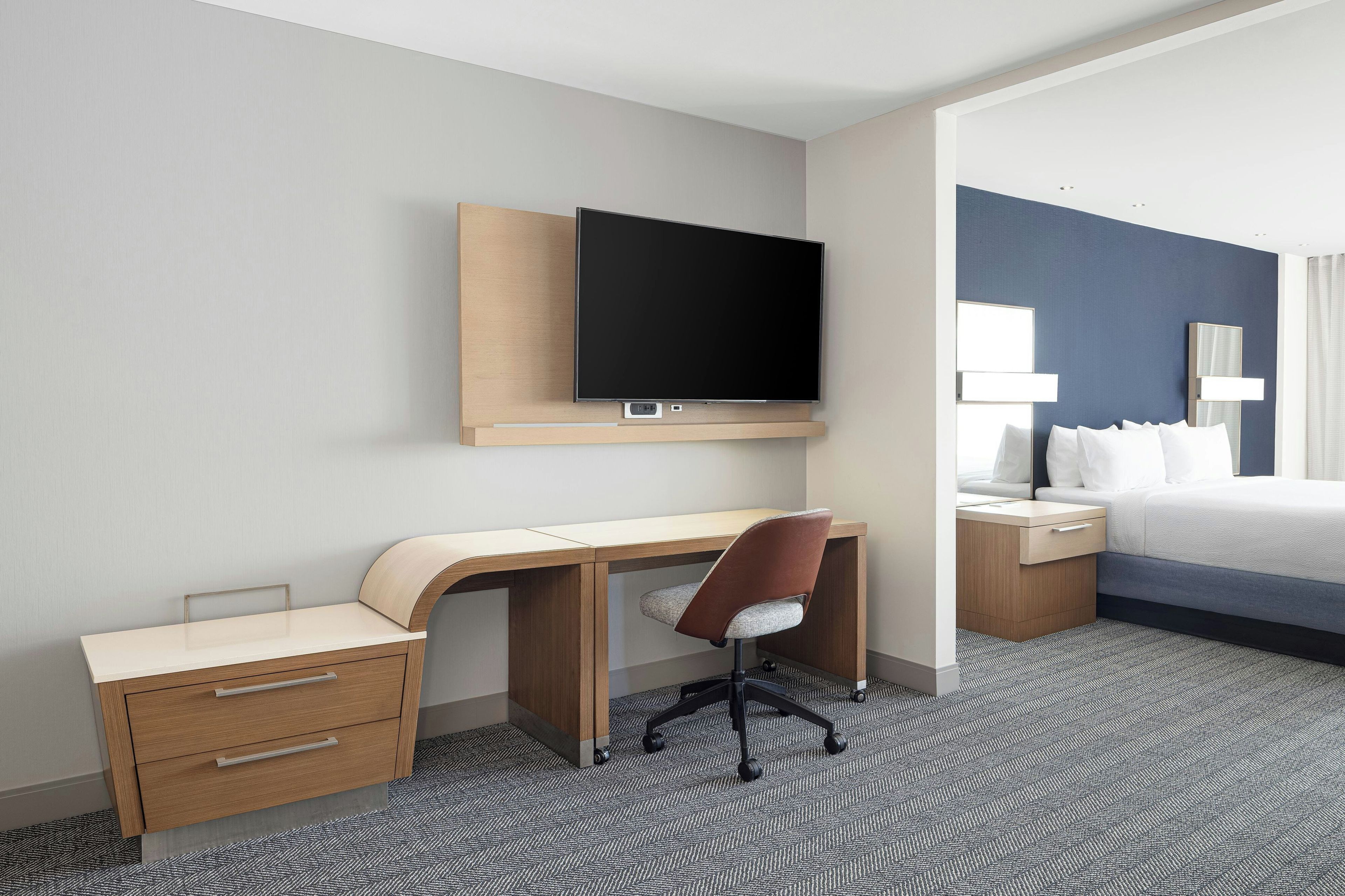 Quarto de hotel com escrivaninha, cadeira, cama, mesas de cabeceira e televisão.