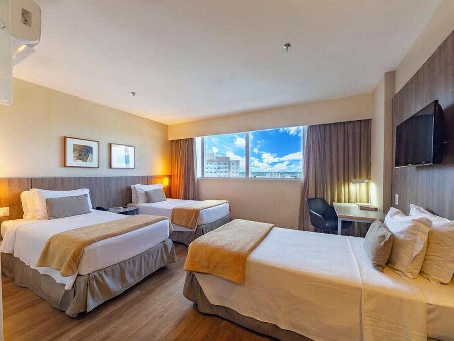 Quarto de hotel com duas camas, uma escrivaninha, uma cadeira, uma televisão e uma janela vista para a cidade.