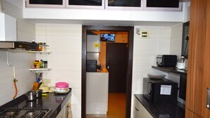 Uma cozinha compacta com fogão, micro-ondas e diversos eletrodomésticos. Há uma geladeira pequena e uma televisão fixada na parede.