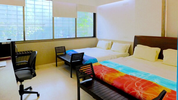 Quarto de hotel com cama, duas cadeiras, escrivaninha e janelas amplas.