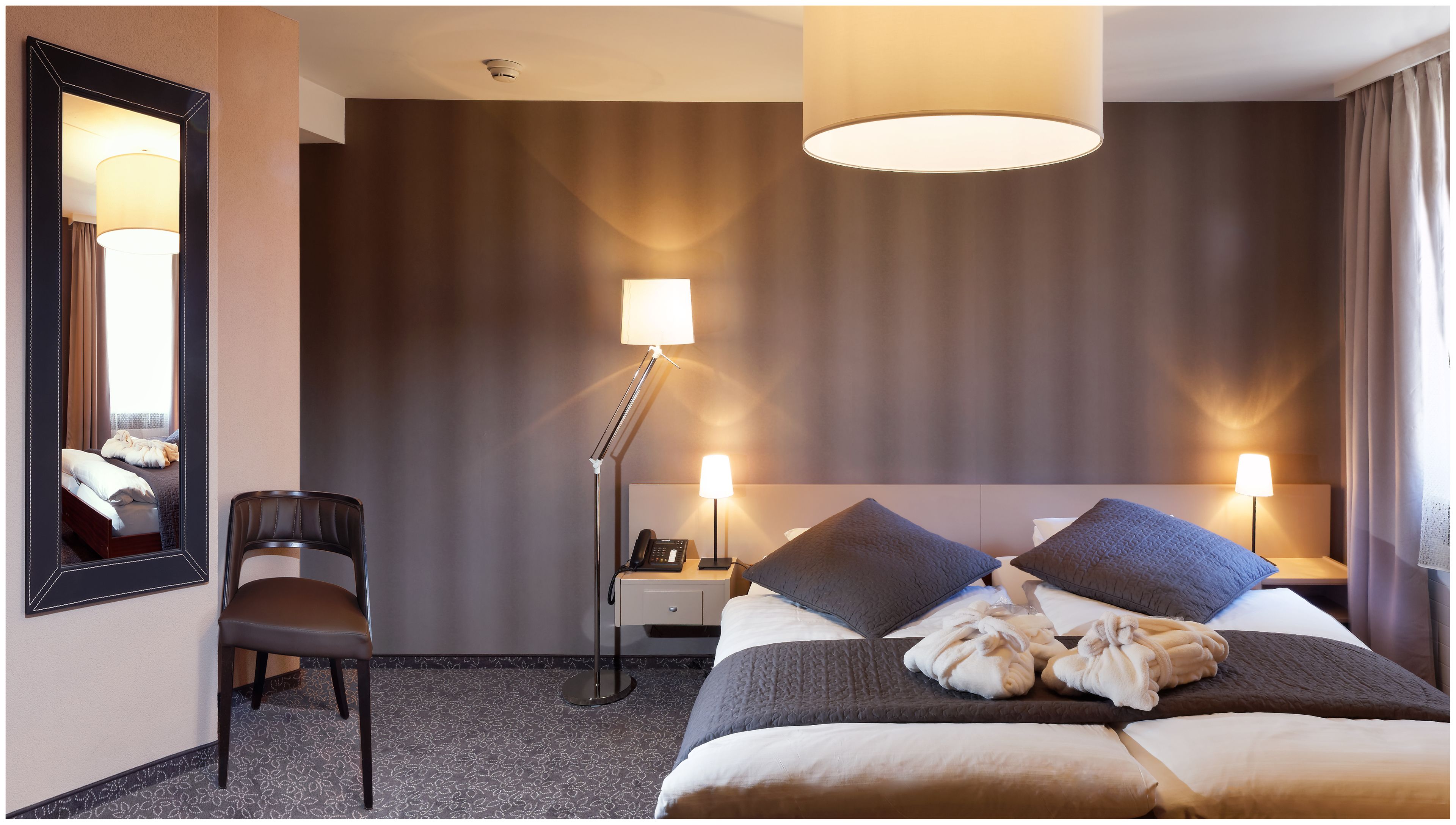 Quarto de hotel com uma cama grande, duas luminárias de cabeceira, uma cadeira, mesa de cabeceira com telefone e um espelho na parede.