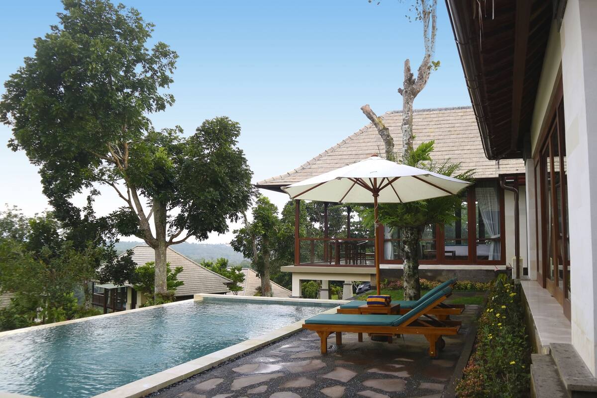 Piscina com espreguiçadeiras e um guarda-sol, cercada por árvores e um edifício com grandes janelas.