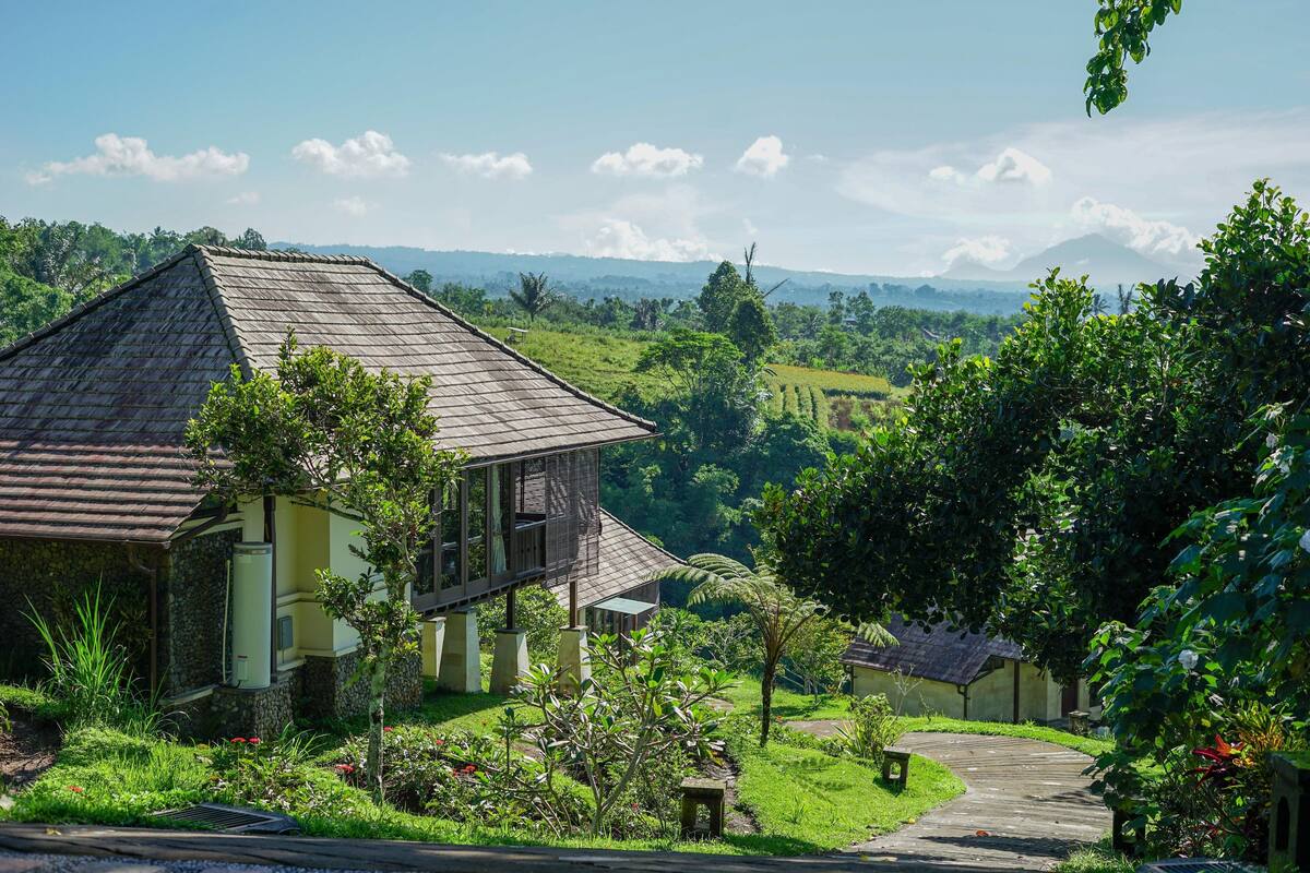 Uma casa tradicional com telhado inclinado, rodeada por vegetação exuberante e vista clara para montanhas distantes.