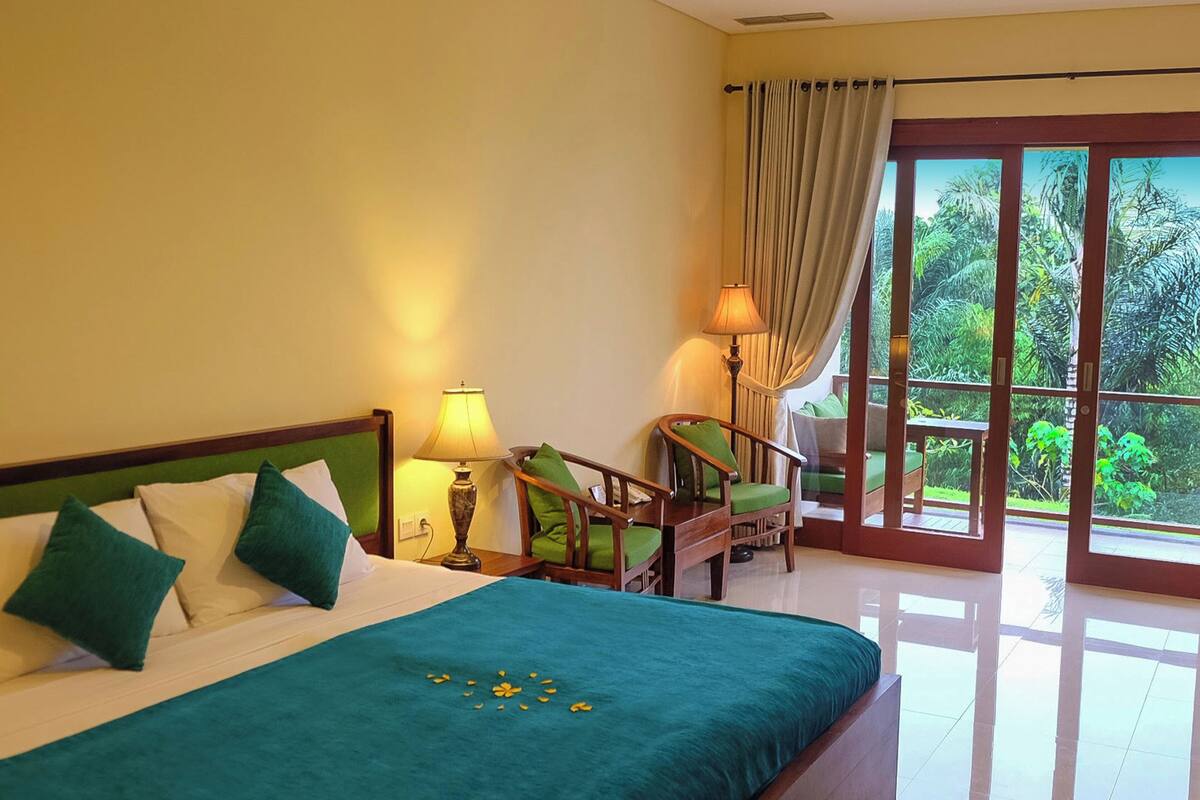 Quarto de hotel com cama, duas cadeiras, uma mesa, um abajur e vista para o verde através de grandes janelas.
