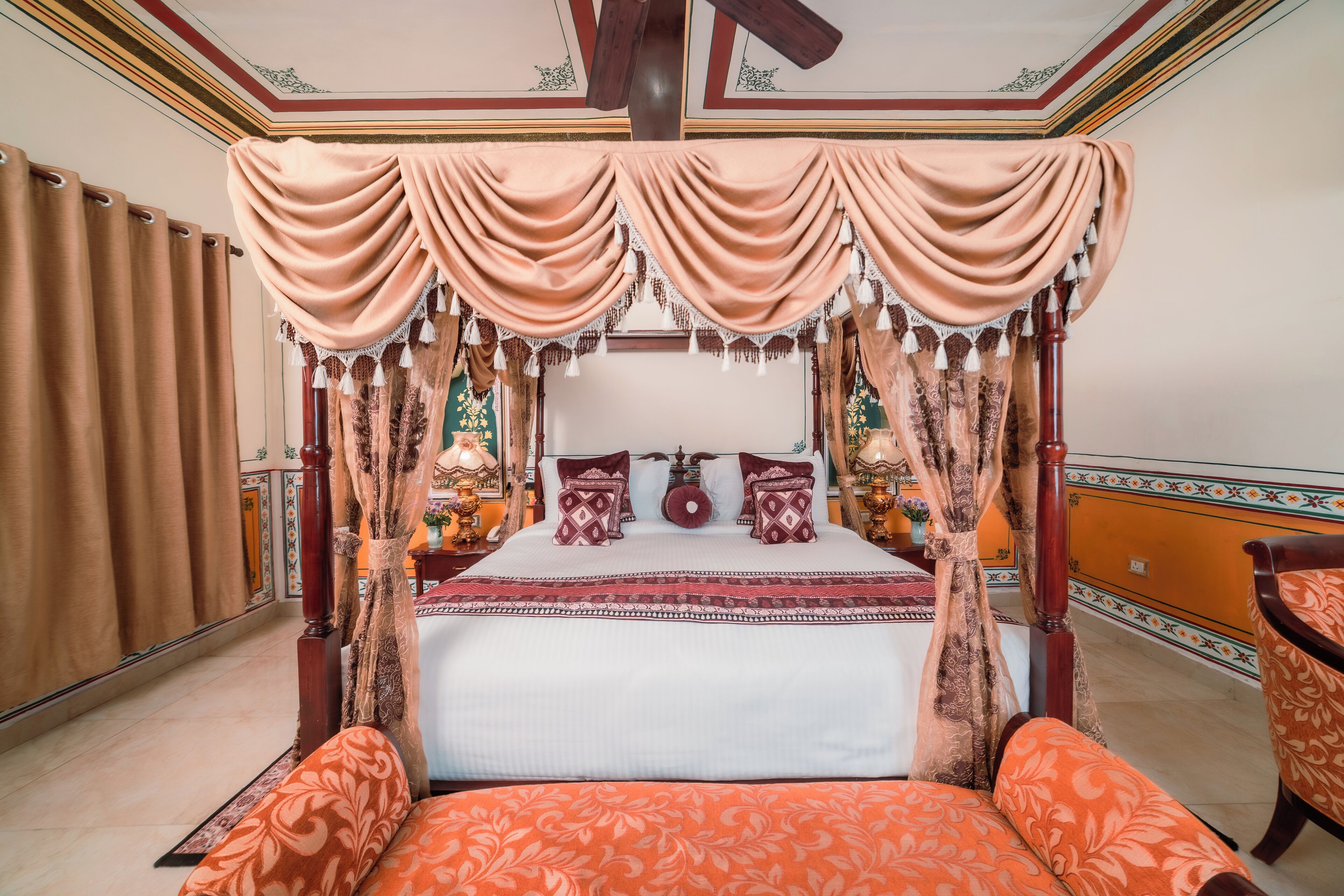 Uma cama com dossel e cortinas elaboradas, um sofá estampado com flores e uma cabeceira decorativa.