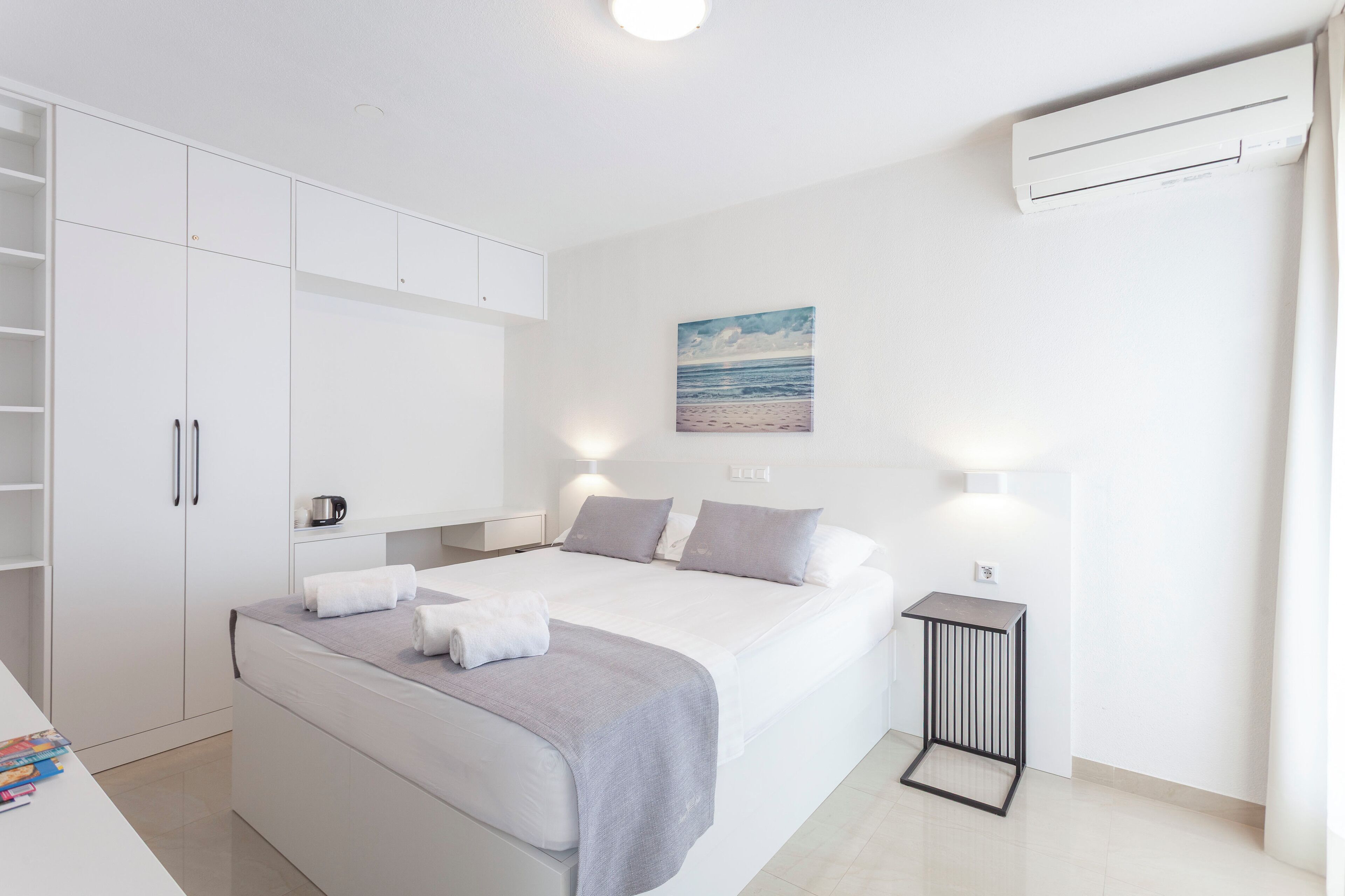 Um quarto moderno com uma cama grande, paredes brancas, um quadro com tema de praia e uma pequena mesa de cabeceira.