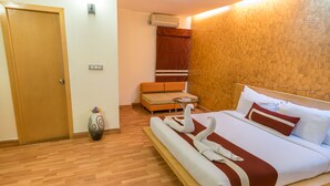 Quarto de hotel com piso de madeira, cama com roupa de cama branca e vermelha, um pequeno sofá e um vaso decorativo.