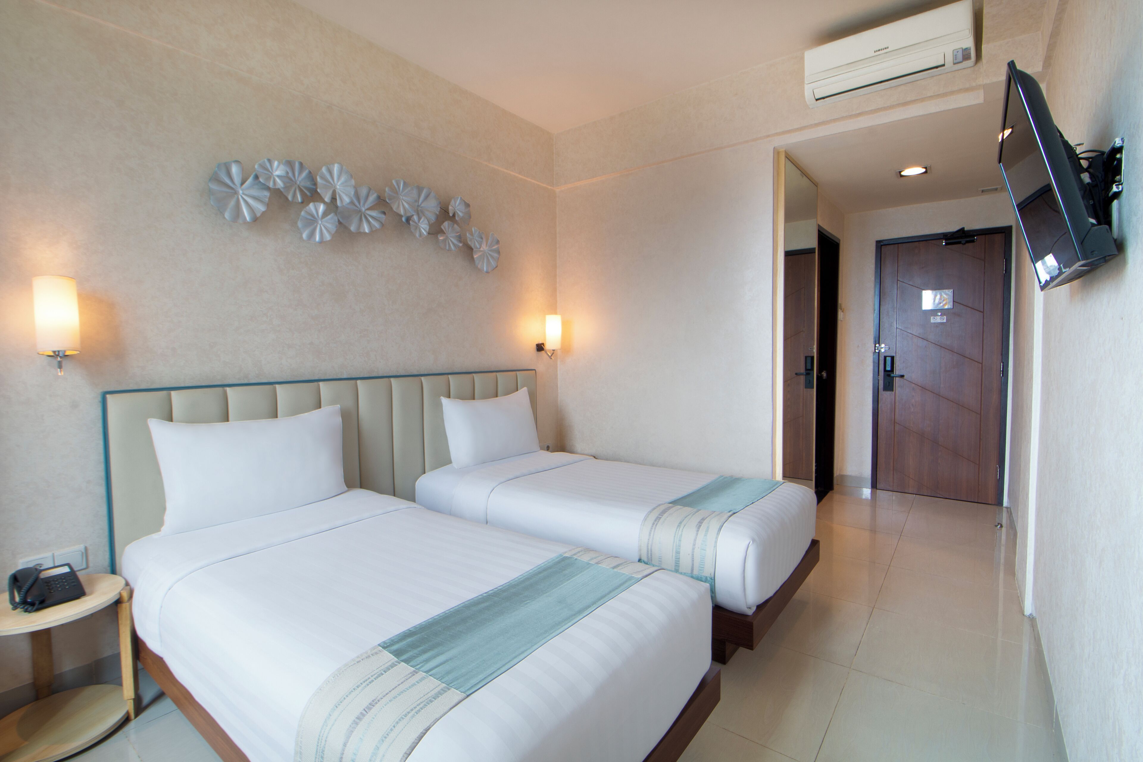 Quarto de hotel com duas camas, mesa de cabeceira com telefone, luminárias na parede e televisão.