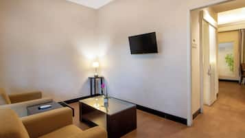 Quarto de hotel com sofá, mesa de centro, televisão de tela plana na parede e abajur em uma mesinha lateral.
