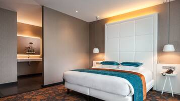 Quarto de hotel moderno com uma cama grande, uma mesa de cabeceira com abajur, nicho embutido com produtos de higiene pessoal e telefone fixo na parede.