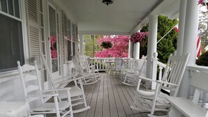 Varanda coberta com cadeiras de balanço brancas e uma mesa central, cercada por janelas e vista para flores cor-de-rosa.