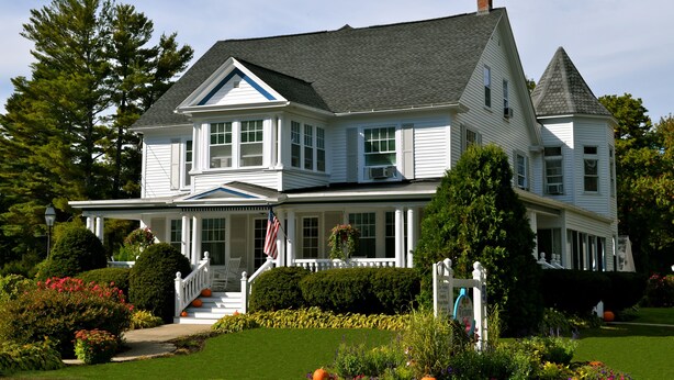 Uma casa grande de dois andares com varanda em volta, chaminé e um jardim bem cuidado com diversas plantas e flores.