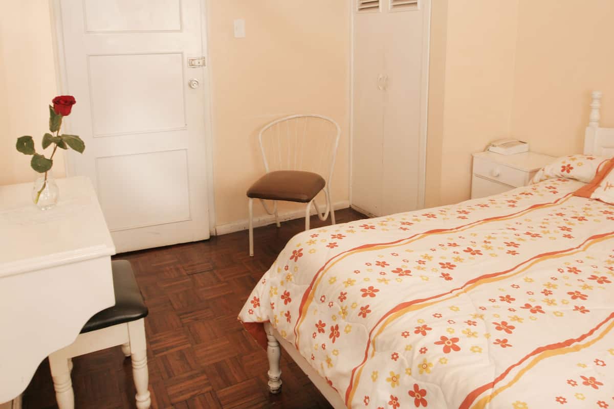 Quarto com cama, poltrona, mesa de cabeceira, cômoda e um vaso com uma rosa.