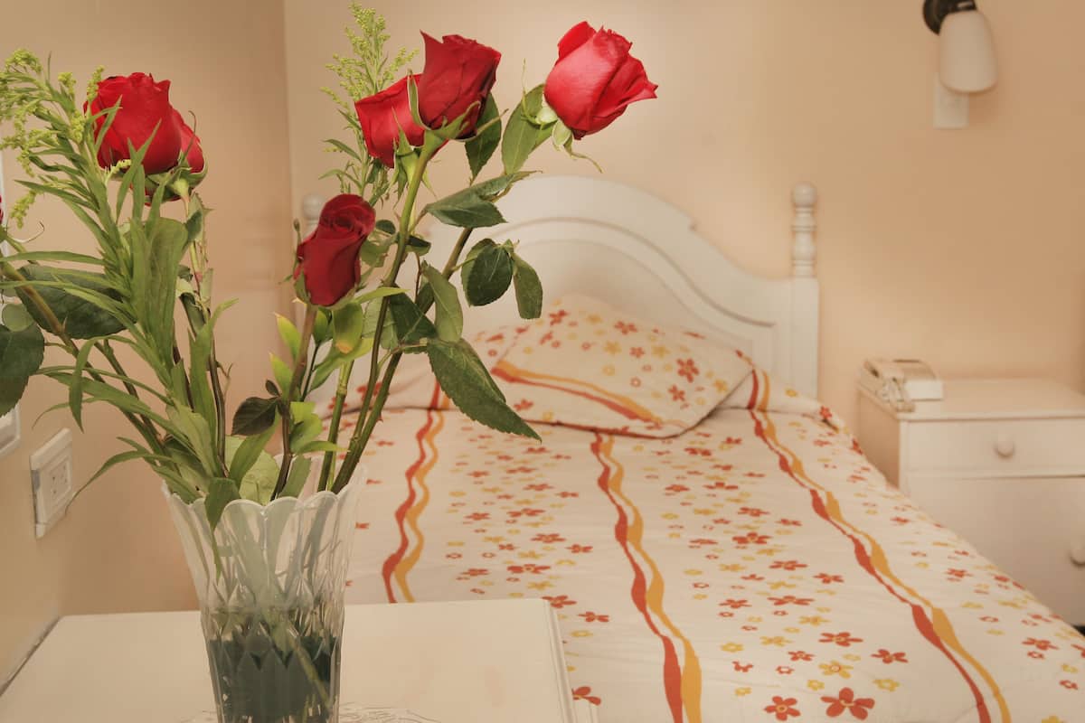 Uma cama com colcha florida, uma mesa de cabeceira e um vaso com rosas vermelhas.