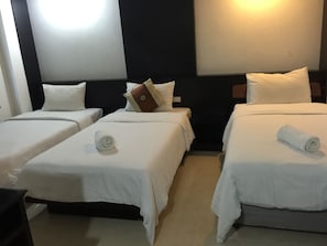 Quarto de hotel com duas camas de solteiro, cada uma com um edredom branco e uma toalha enrolada. O quarto possui uma cabeceira escura e uma parede clara com um aparelho de iluminação.