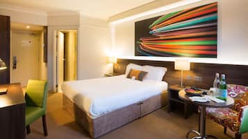 Quarto de hotel com cama, escrivaninha, cadeira, mesa com garrafas e uma obra de arte colorida na parede.