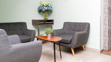 Um quarto com duas poltronas cinzas, uma pequena mesa de madeira com uma planta em vaso, e um vaso com flores azuis sobre uma mesinha próxima a uma parede verde.