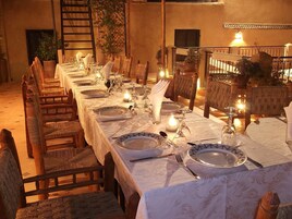 Uma longa mesa de jantar posta com pratos, talheres e velas, rodeada por cadeiras de vime em ambiente externo.