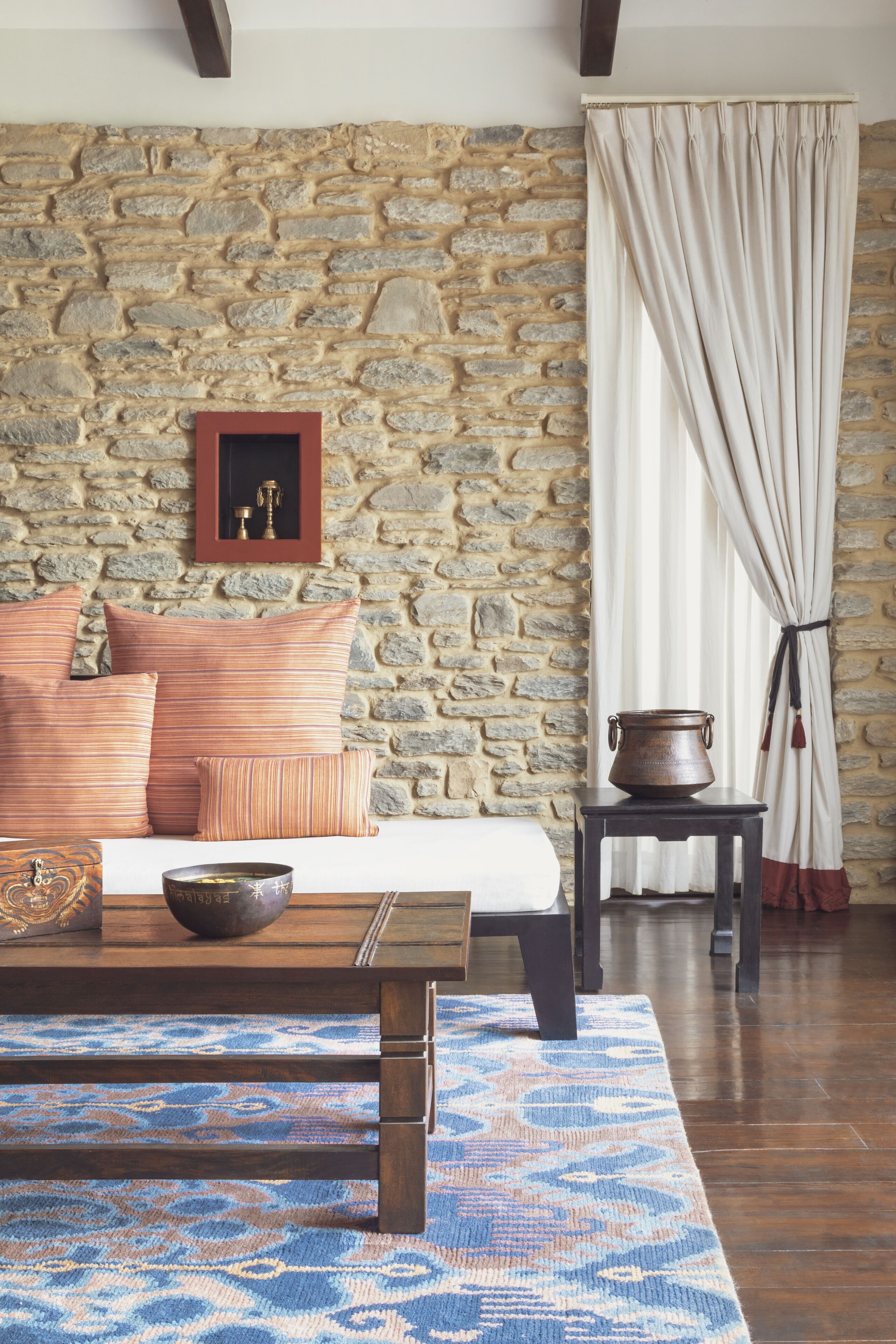 Um quarto com parede de pedra, cama com almofadas listradas, mesa de madeira com uma tigela e cortina.