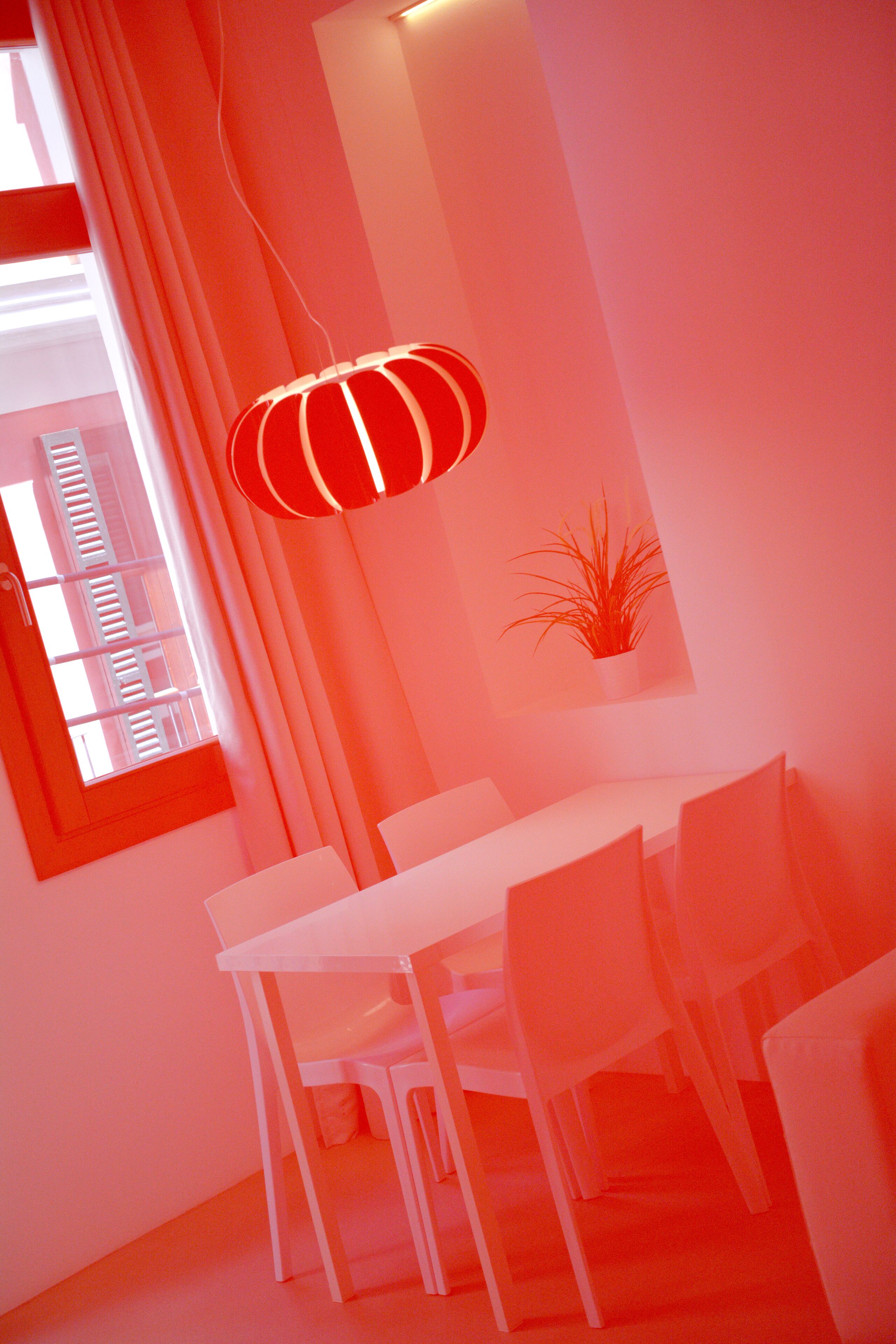 Uma sala de jantar com uma mesa e cadeiras brancas, uma luminária pendente vermelha e um vaso de planta na parede.