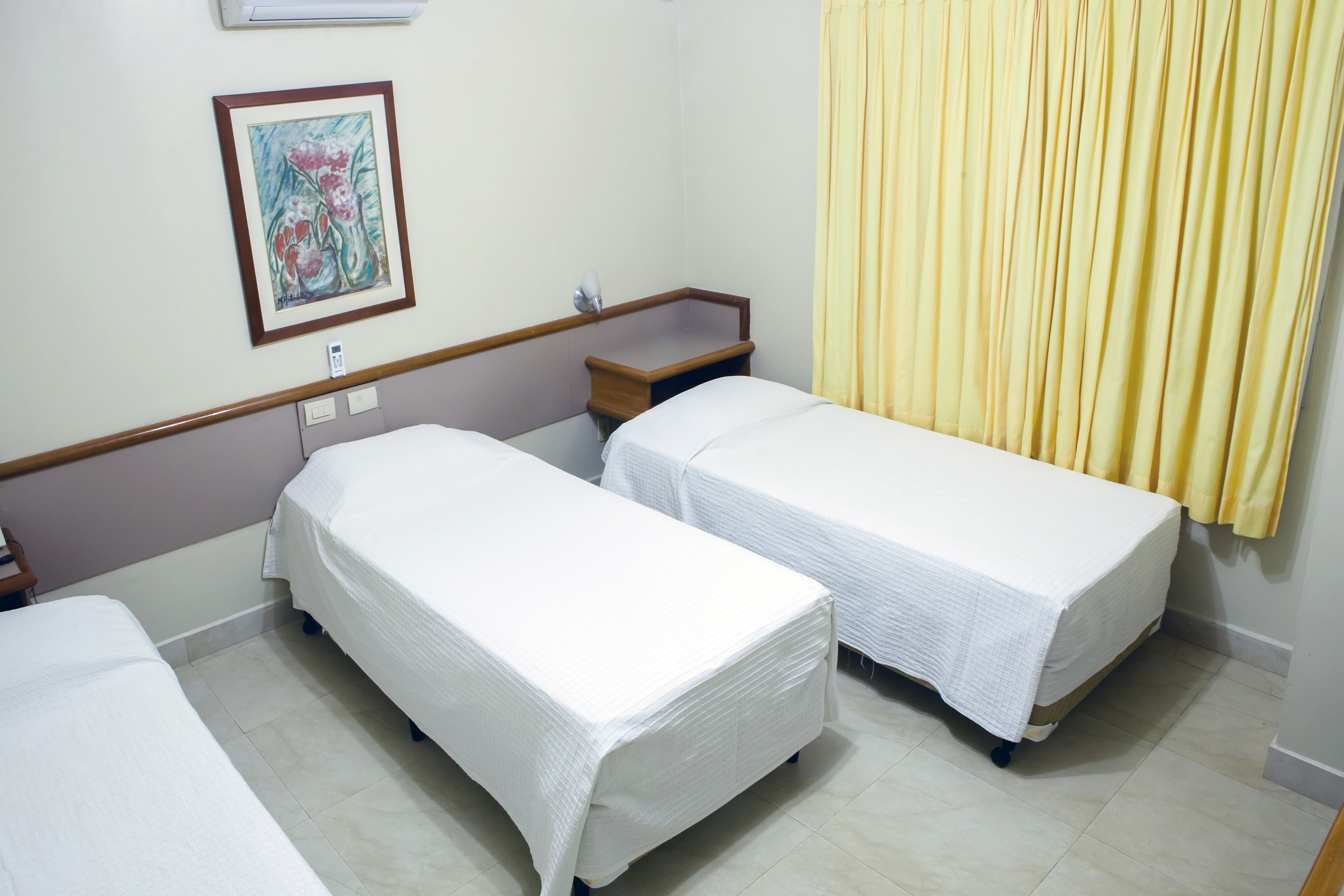 Quarto de hotel com duas camas de solteiro, uma mesinha de cabeceira, uma luminária e um quadro na parede, com cortina amarela.