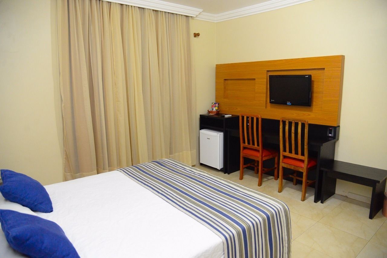 Quarto de hotel com cama, escrivaninha, televisão e um pequeno frigorífico.