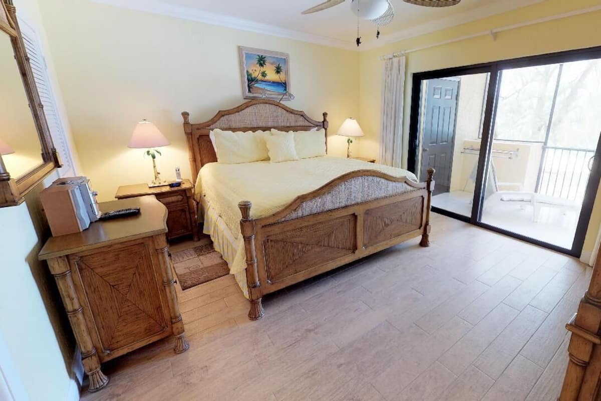 Quarto com cama de madeira, mesas de cabeceira, ventilador de teto e porta de vidro deslizante para uma varanda.