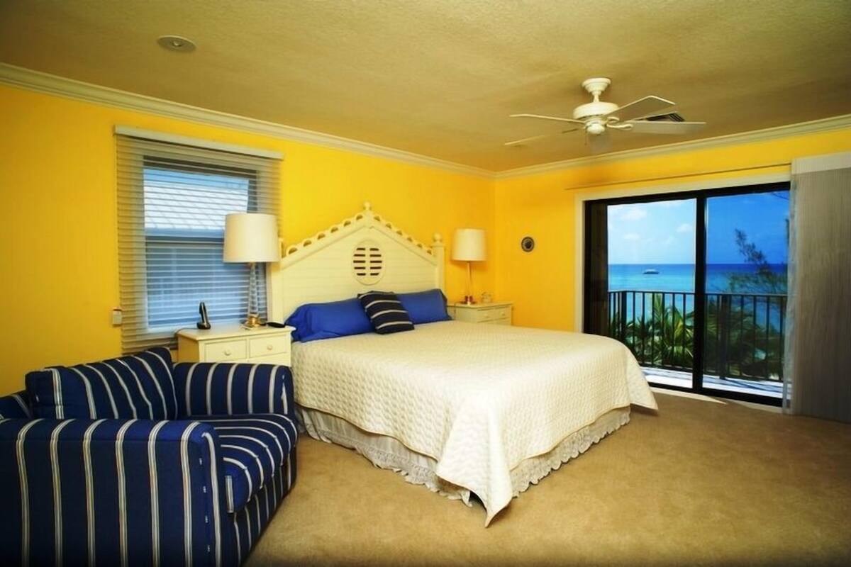 Quarto com cama, sofá, ventilador de teto e vista para o oceano.