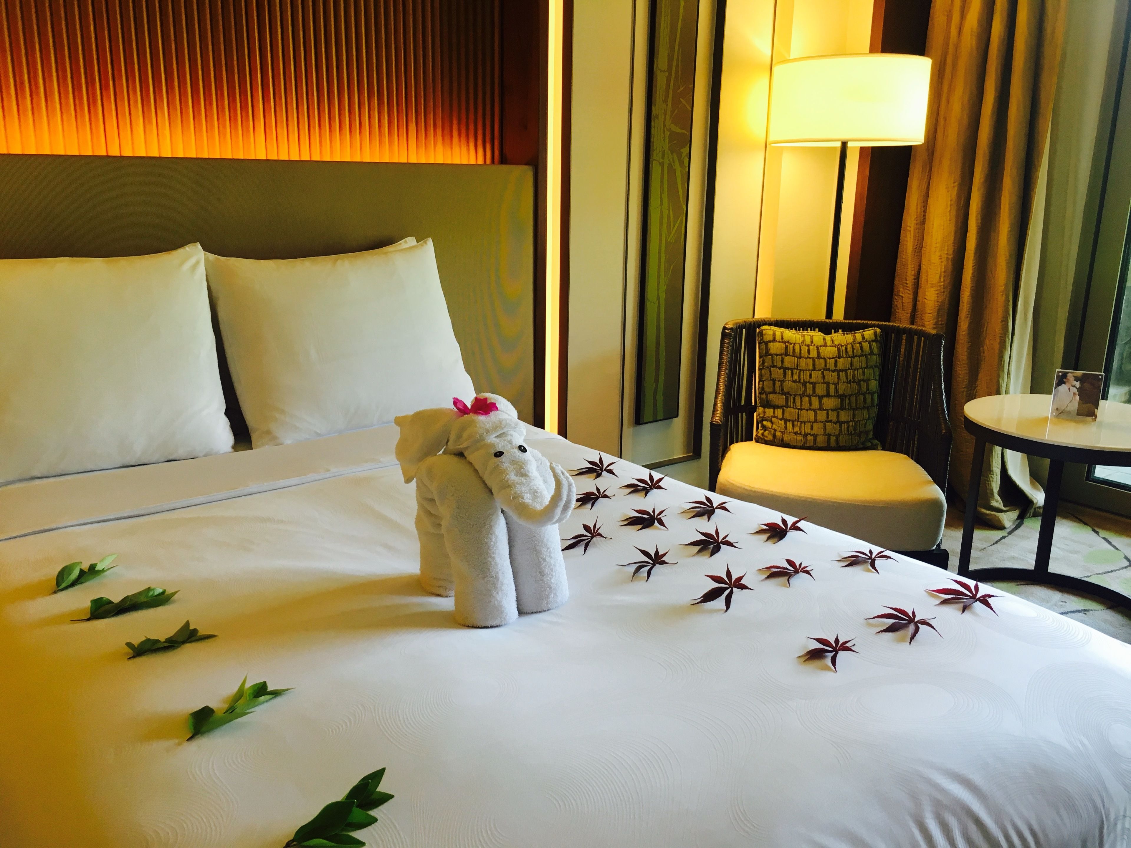 Quarto de hotel com cama bem arrumada, poltrona confortável, pequena mesa e luminária.