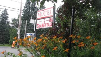 Placa de motel com esquema de cores vermelho e branco, rodeada por vegetação exuberante e flores laranjas.