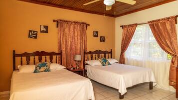 Quarto de hotel com duas camas de solteiro, cabeceiras de madeira, um ventilador de teto e uma janela com cortinas.