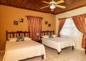 Quarto de hotel com duas camas de solteiro, cabeceiras de madeira, um ventilador de teto e uma janela com cortinas.