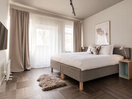 Quarto com cama, cortinas, tapete, mesa de cabeceira e luminária fixada na parede.