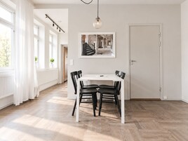 Uma área de refeições com mesa branca e cadeiras pretas, um quadro na parede e uma porta à direita.