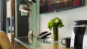 Quarto de hotel com escrivaninha de vidro, telefone, cafeteira, um vaso com flores e vista para a cidade na parede.