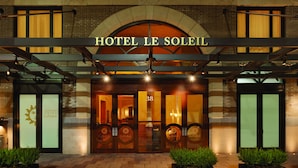 A entrada do Hotel Le Soleil, com fachada de tijolo, grandes portas de vidro e uma placa sobre a entrada.