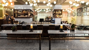 Um restaurante com várias mesas e cadeiras, um bar com prateleiras cheias de garrafas, e uma parede decorada com quadros.