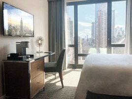 Quarto de hotel com uma cama, uma escrivaninha com cafeteira, uma cadeira e vista da cidade através de grandes janelas, além de um quadro com paisagem urbana.