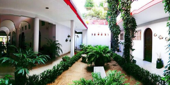 Um pátio com plantas em vasos, uma área central para sentar e um edifício branco com arcos.