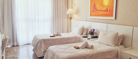 Quarto de hotel com duas camas, mesa de cabeceira com abajur, pintura na parede e uma janela grande com cortinas.
