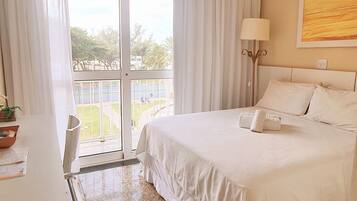 Quarto de hotel com cama, escrivaninha, abajur e vista para uma quadra de tênis pela porta.