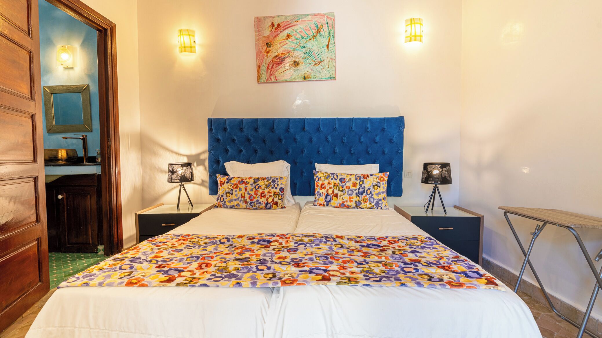 Quarto com cama grande, duas mesas de cabeceira, cabeceira estofada azul e uma pintura abstrata colorida sobre a cama.